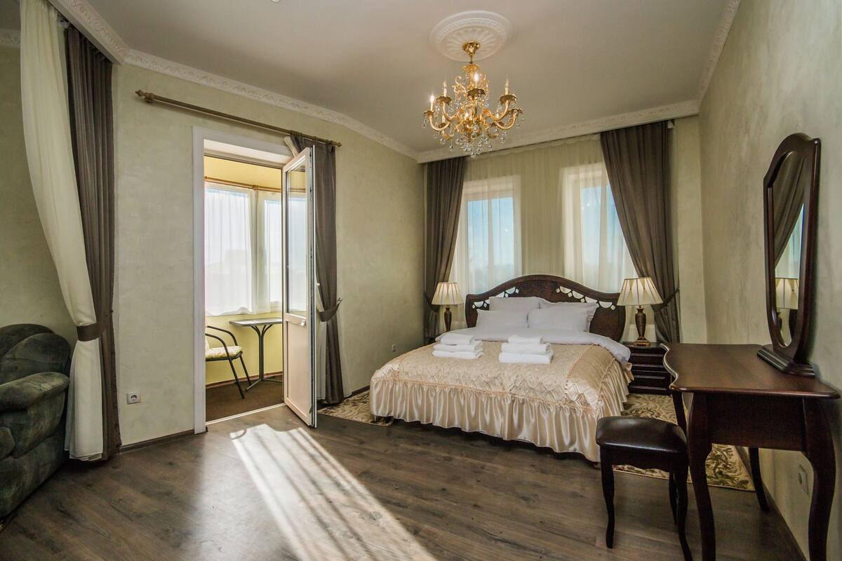 Quarto de hotel com uma cama grande, uma escrivaninha, uma cadeira, um candelabro e vista para o exterior.