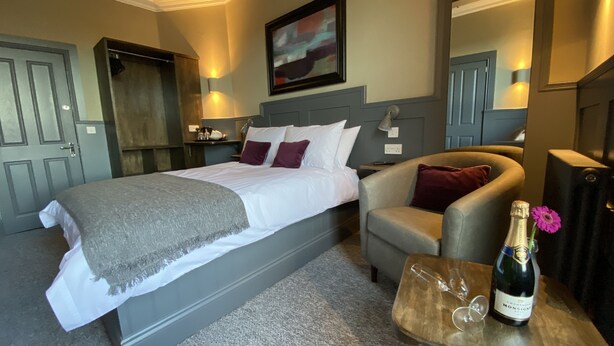 Quarto de hotel com uma cama, uma cadeira, uma mesinha com uma garrafa de champanhe e um espelho.