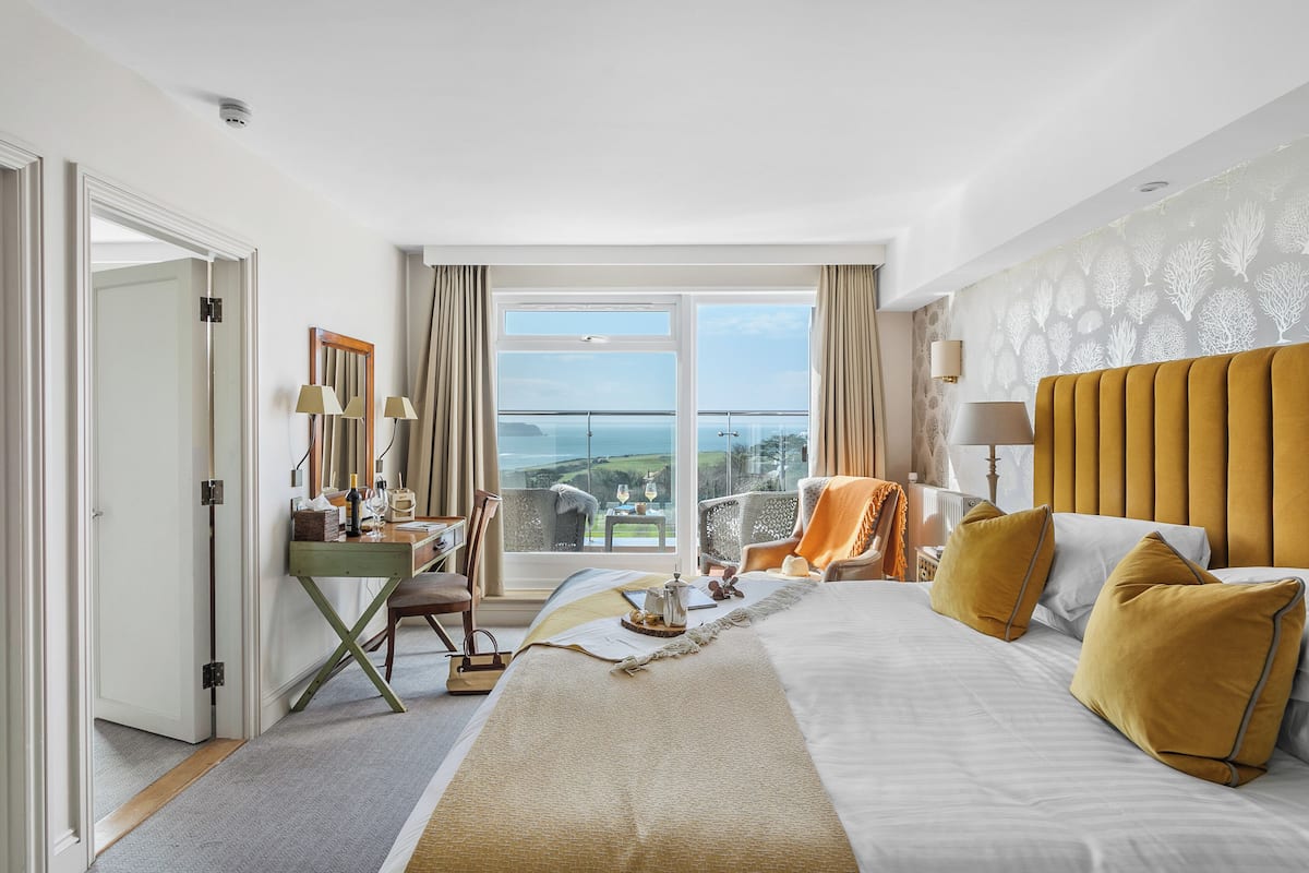Quarto de hotel com uma cama grande, uma escrivaninha com cadeira, vista para o mar e uma varanda com mesa e cadeiras.