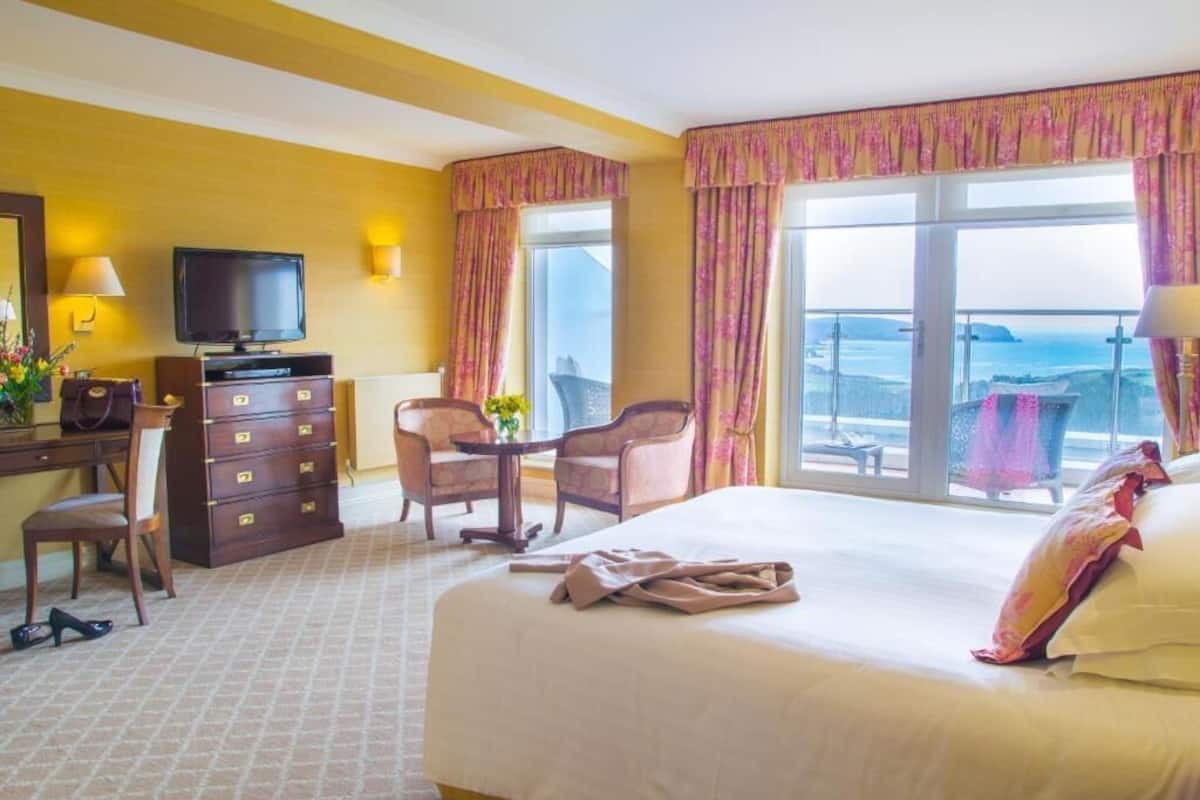 Quarto de hotel com cama, escrivaninha com televisão, cadeira, cômoda e varanda com vista.