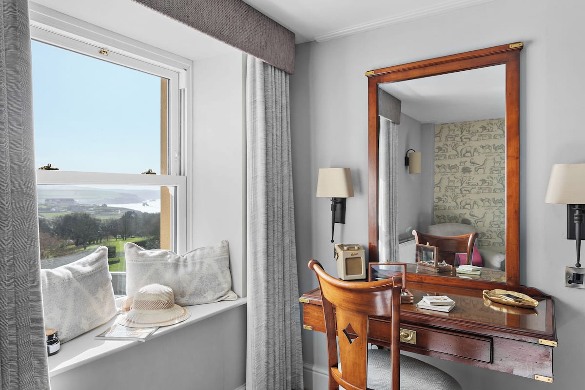 Quarto de hotel com uma grande janela, uma escrivaninha de madeira, um espelho e vista para o campo.