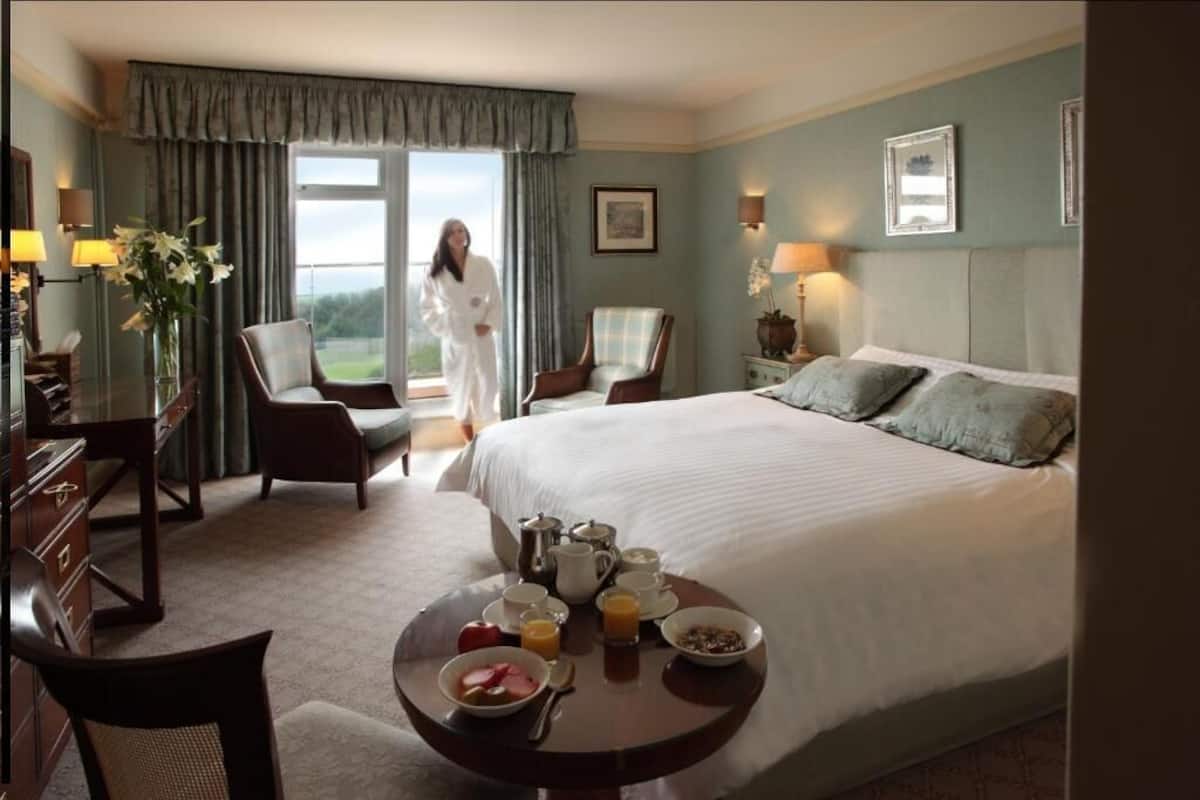 Quarto de hotel com uma cama grande, uma mesa de jantar com café da manhã, um espaço de estar com cadeiras e vista para o exterior.