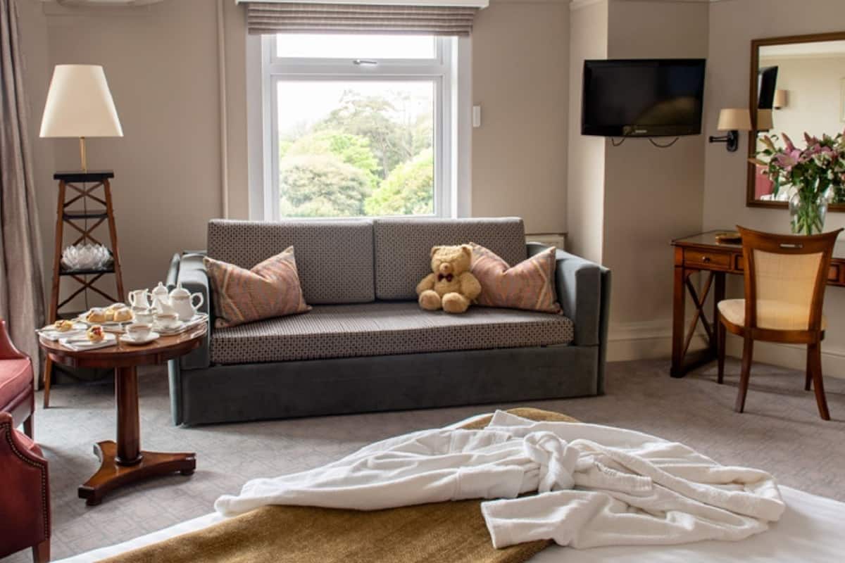 Quarto de hotel com poltrona vermelha, sofá cinzento com almofadas, mesa pequena com serviço de chá, mesa de cabeceira com abajur, televisão e espelho.