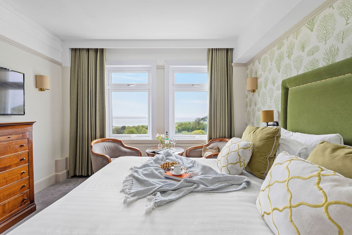 Quarto de hotel com cama grande, penteadeira, televisão, janela com cortinas e vista para o verde lá fora.