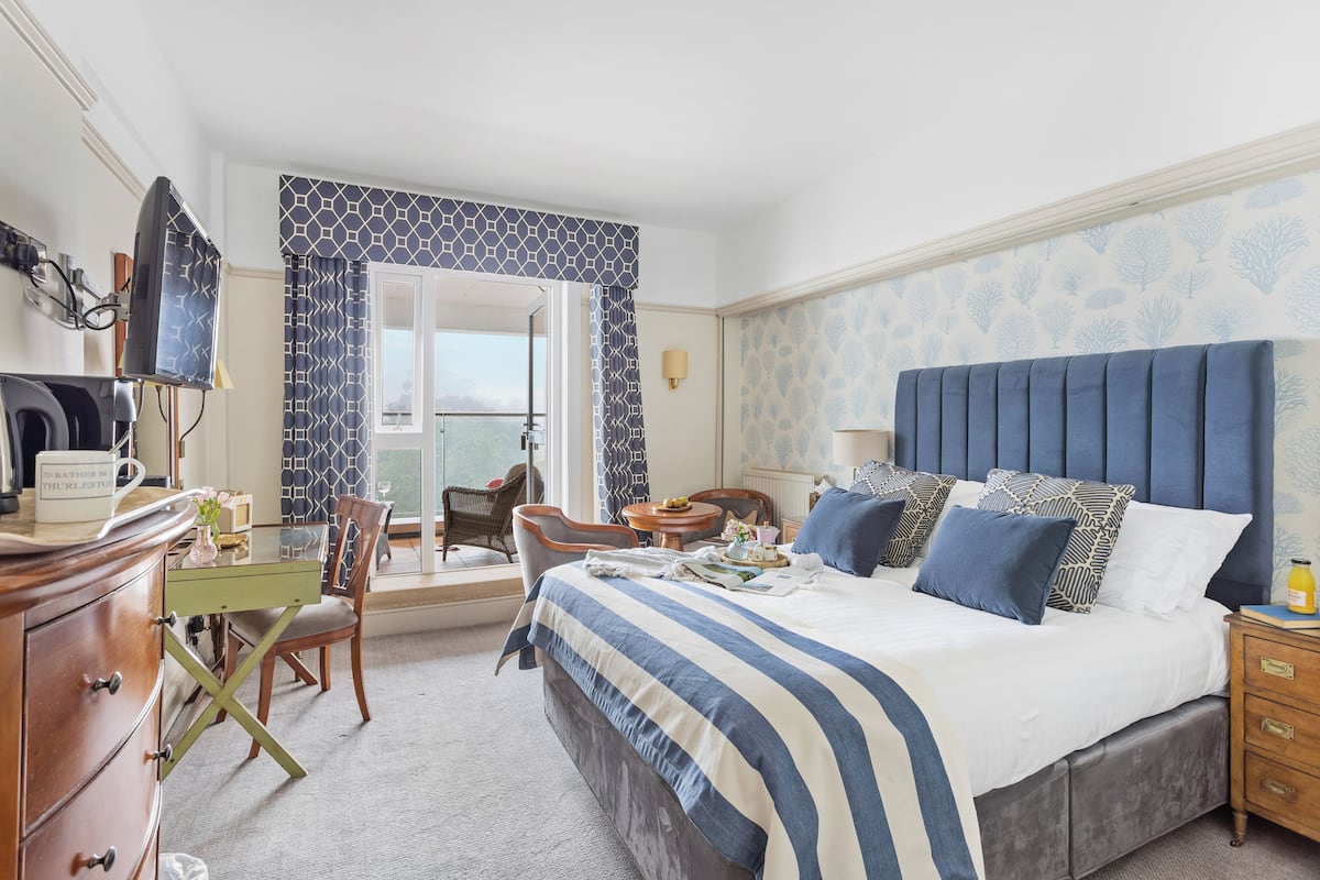 Quarto de hotel com cama, escrivaninha, cadeira, cômoda e janela com cortinas.