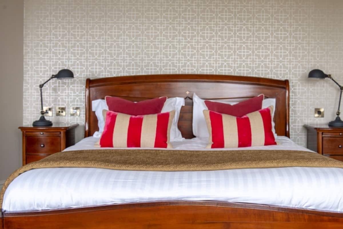 Uma cama bem arrumada com cabeceira de madeira, duas mesas de cabeceira com abajures e papel de parede estampado.