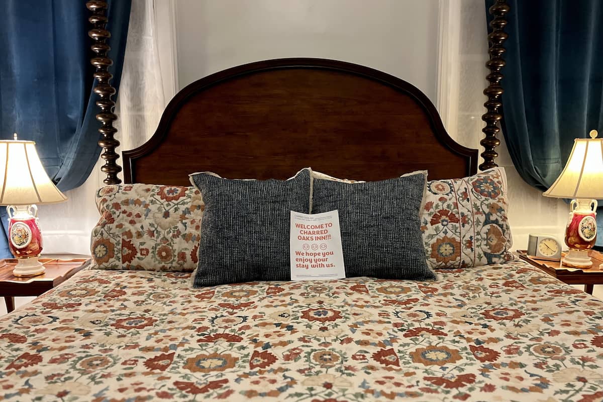Uma cama com colcha estampada com flores, cabeceira escura e duas almofadas decorativas. Um letreiro de boas-vindas está colocado sobre a cama.