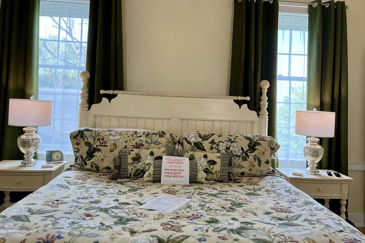 Cama arrumada com roupa de cama florida, duas luminárias de cabeceira, uma placa de boas-vindas e vista para o exterior através de uma janela com cortinas.