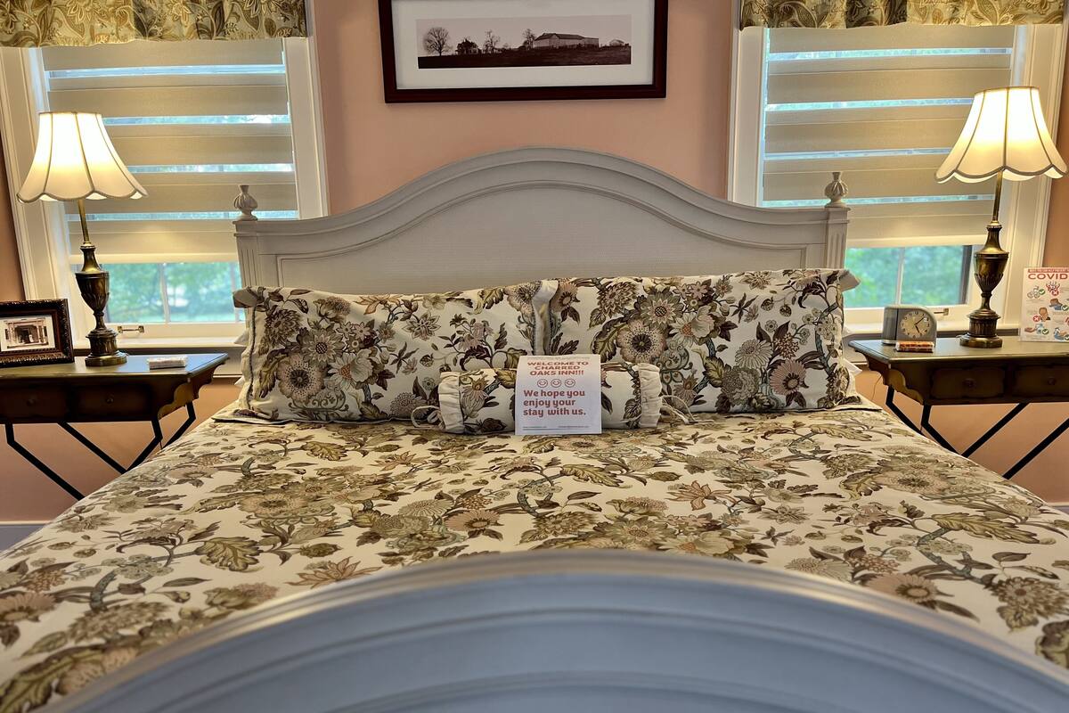 Cama bem arrumada com roupa de cama florida, duas mesas de cabeceira com luminárias, um quadro na mesa da esquerda e um sinal de boas-vindas na cama.