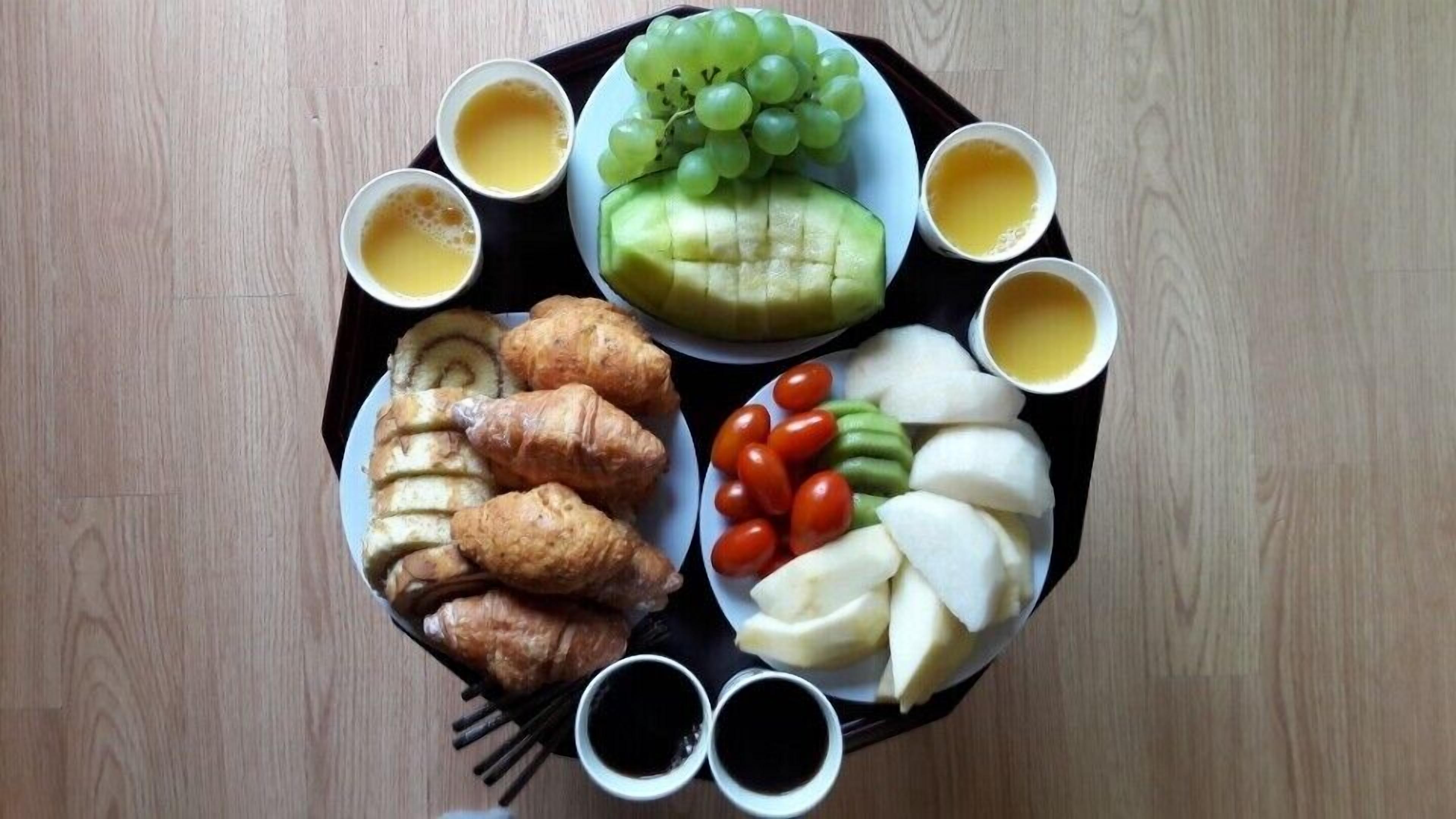 Uma bandeja de refeição com diversos alimentos, incluindo uvas, melão, croissants, tomates cereja e dois tipos de molhos.