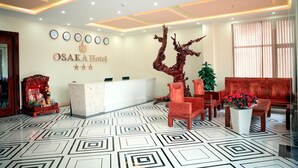 Uma área de recepção de hotel com design moderno, apresentando um balcão de atendimento, espaço de estar, escultura de árvore decorativa e piso estampado.