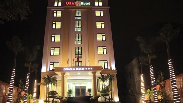 Um hotel com vários andares, sinalização iluminada, entrada central com degraus e plantas em vasos ao redor da entrada.