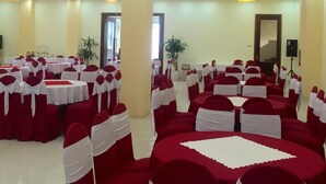 Salão de banquetes com mesas redondas e cadeiras, decoração em tons de branco e vermelho, e iluminação embutida.