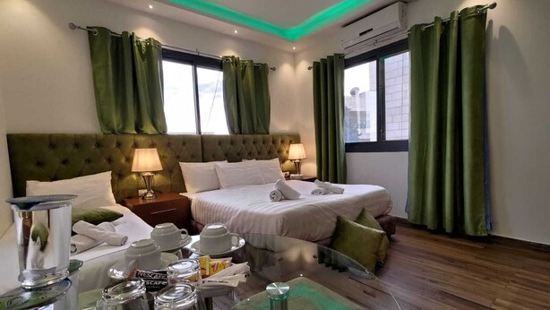 Quarto de hotel com uma cama grande, duas mesas de cabeceira, uma escrivaninha e uma cadeira. Há cortinas, uma janela e um aparelho de ar condicionado.