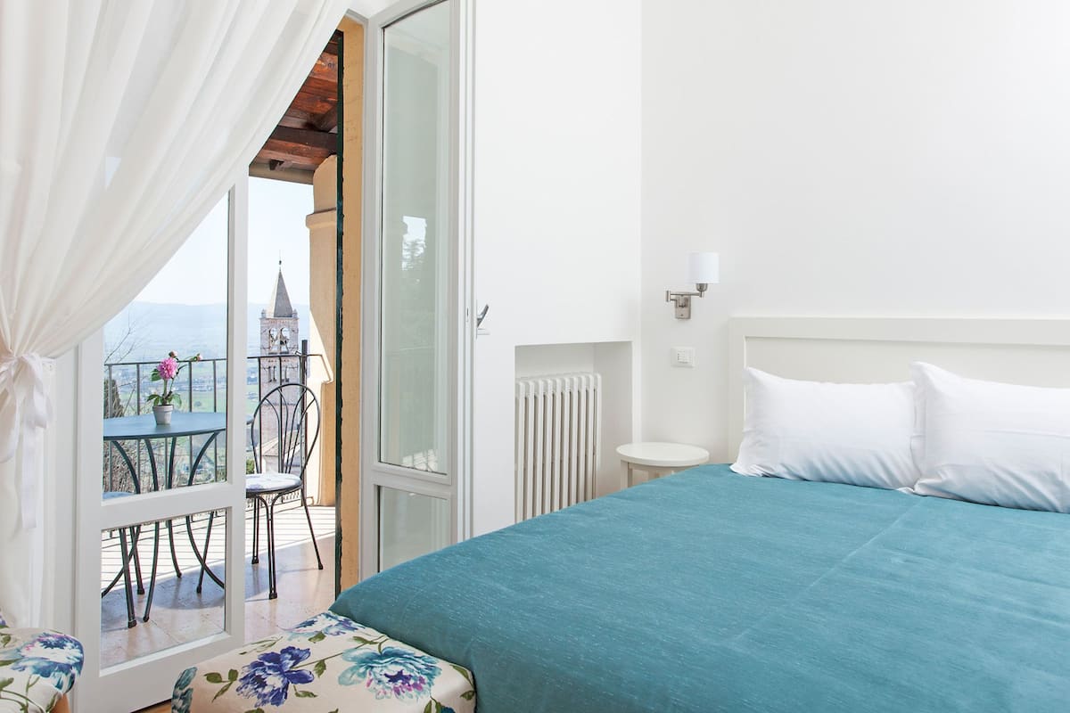 Quarto com cama grande, vista de uma torre através de uma porta de varanda, mesa pequena com cadeiras e radiador.