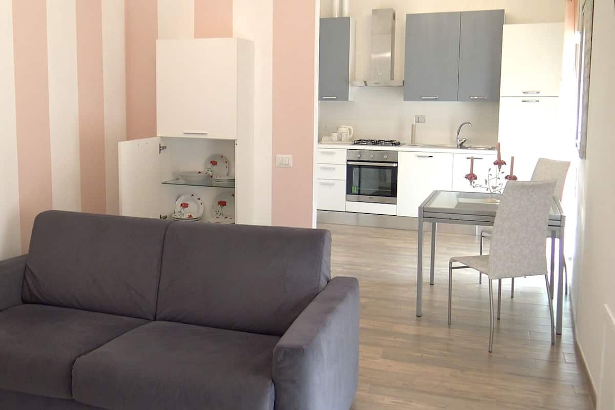 Uma sala de estar moderna com um sofá cinzento, uma área de jantar com mesa e cadeiras de vidro, e uma cozinha com armários brancos e eletrodomésticos de aço inoxidável.