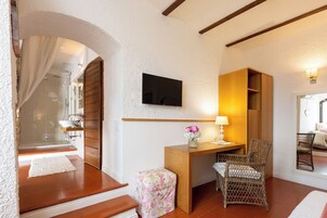 Quarto de hotel com secretária de madeira, cadeiras de vime, televisão na parede, área de chuveiro e cama com vista para o quarto.
