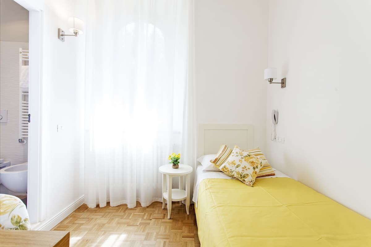Quarto de hotel com cama, uma pequena mesa com um vaso de flores, banheiro com banheira e uma parede branca com luminária.