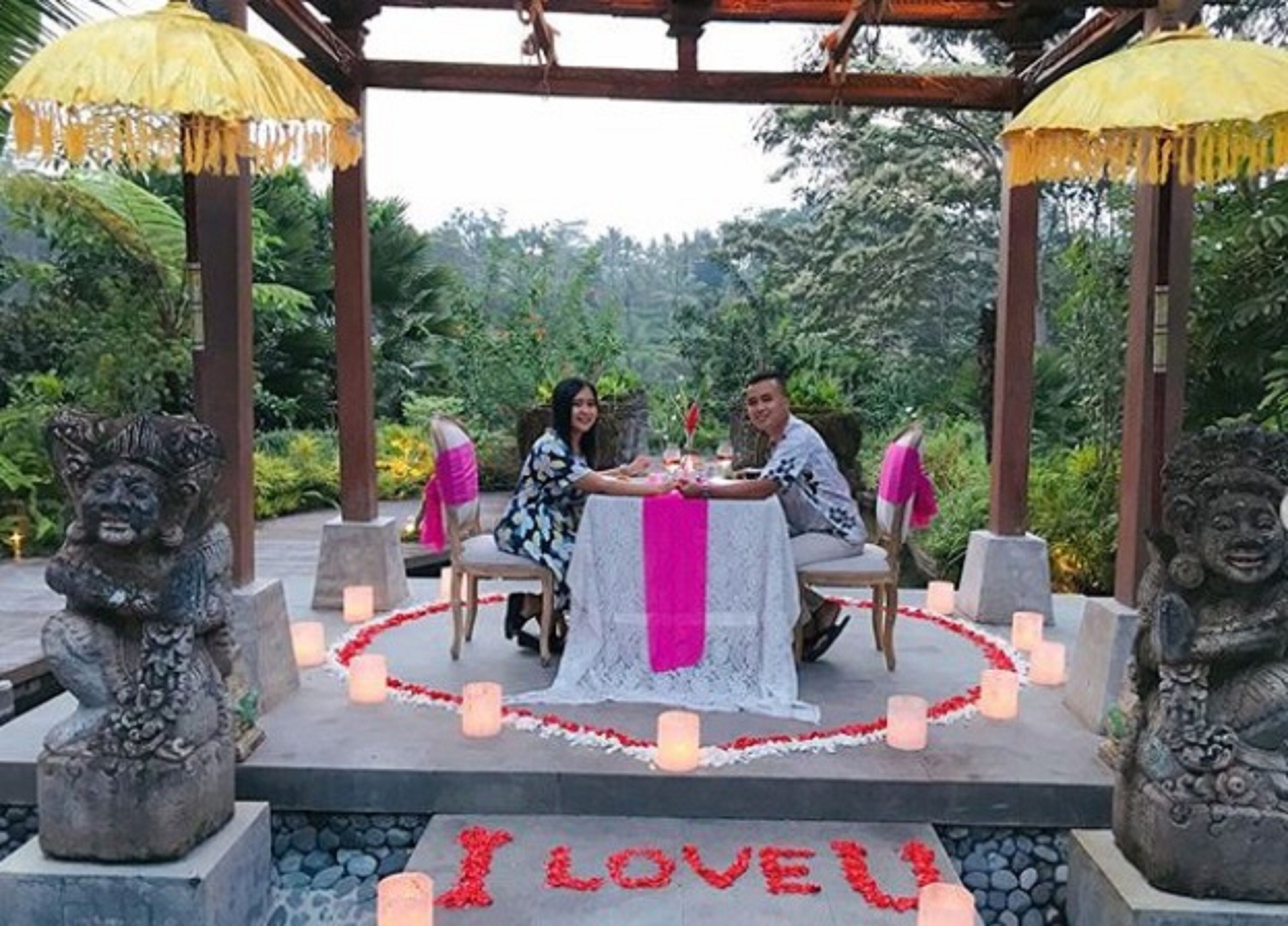 Um casal sentado à mesa com toalha rendada branca, rodeado por velas e a mensagem 'I LOVE YOU' no chão.