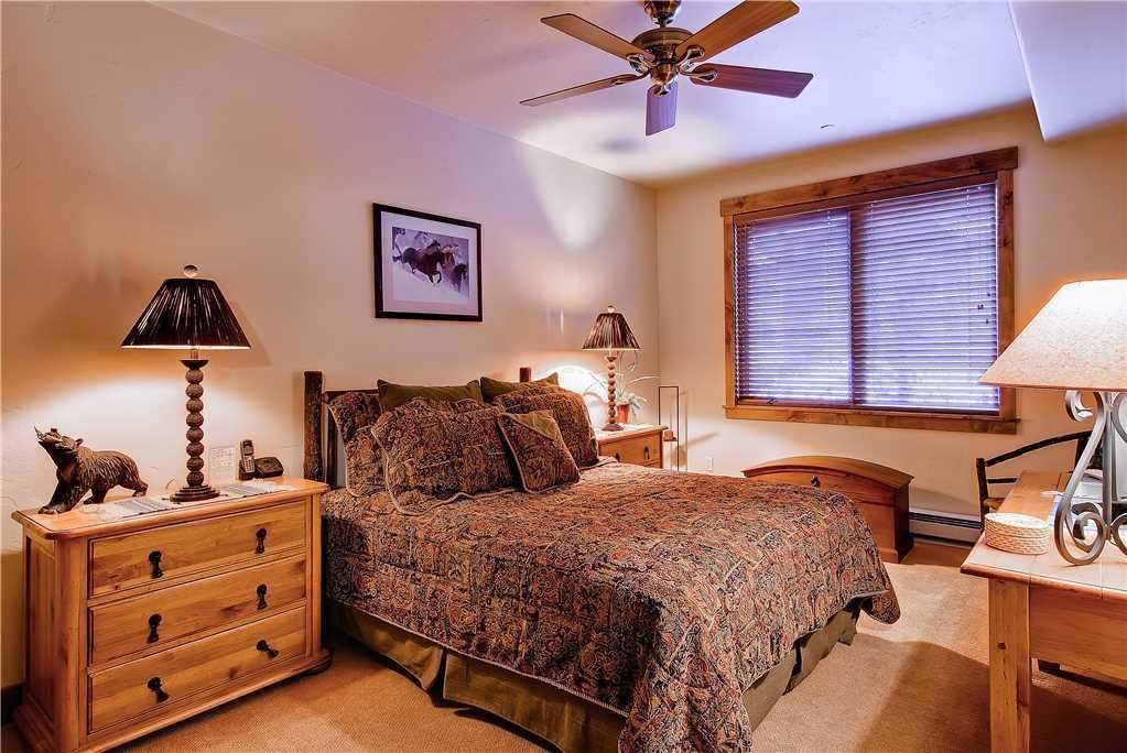 Quarto com cama, mesas de cabeceira de madeira, ventoinha no teto, janela com persianas e quadro pendurado na parede.
