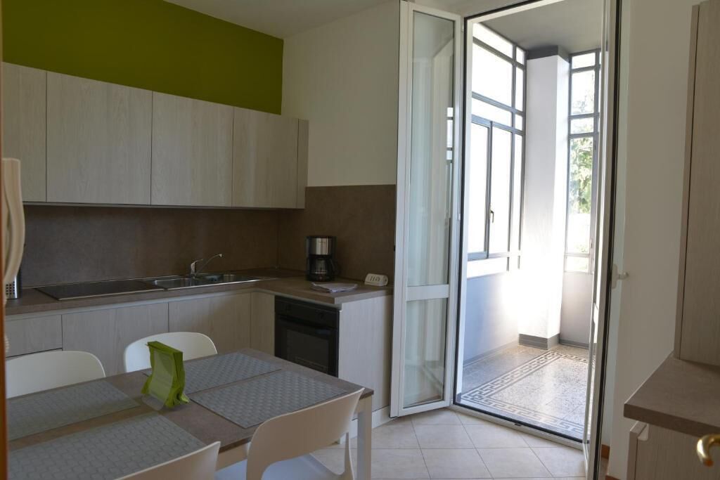 Uma cozinha compacta com pia, fogão e máquina de café. Há uma mesa de jantar com cadeiras e uma porta de vidro deslizante que dá para uma área externa.