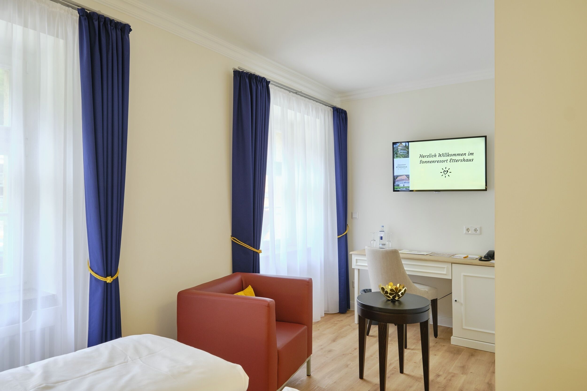 Quarto de hotel com cama, poltrona vermelha, mesinha com abajur, escrivaninha e televisão exibindo uma mensagem de boas-vindas.