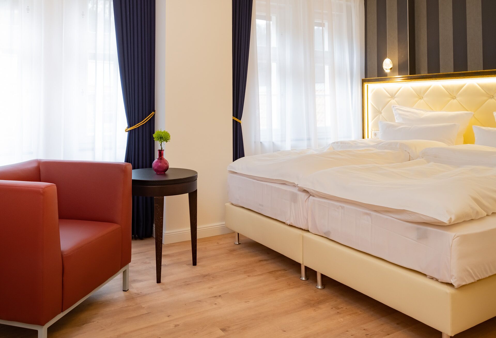 Quarto de hotel com uma cama, uma poltrona vermelha, uma mesinha com um vaso e cortinas leves.