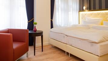 Quarto de hotel com uma cama, uma poltrona vermelha, uma mesinha com um vaso e cortinas leves.