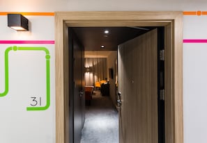 Um corredor de hotel com uma porta de madeira, o número '31' na parede e um quarto ao fundo.