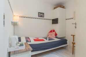 Quarto pequeno e bem organizado, com uma cama, uma escrivaninha com chaleira, uma cadeira e uma TV na parede.