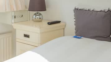 Uma cama bem arrumada com almofada cinza, mesa de cabeceira com abajur e controle remoto, e uma gavetinha.