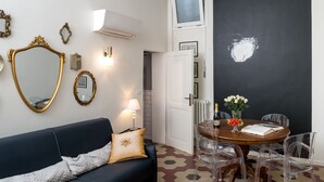 Uma sala de estar moderna com um sofá azul escuro, uma mesa de jantar redonda de madeira e cadeiras transparentes, além de uma parede com lousa.