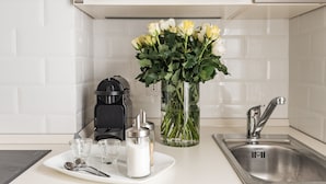 Uma cozinha com pia de aço inoxidável, cafeteira, um vaso de vidro com flores e um prato branco com utensílios.