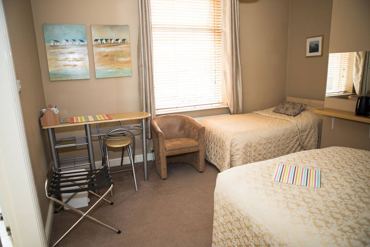Quarto de hotel compacto com uma cama de solteiro, uma pequena escrivaninha com cadeira, uma janela com persianas e duas pinturas penduradas na parede.