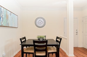 Uma sala de jantar com uma mesa, cadeiras, um relógio na parede e uma porta ao fundo.