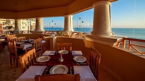 Uma área de refeições com mesas para dois, amplas janelas com vista para o mar e uma varanda com guarda-corpo.