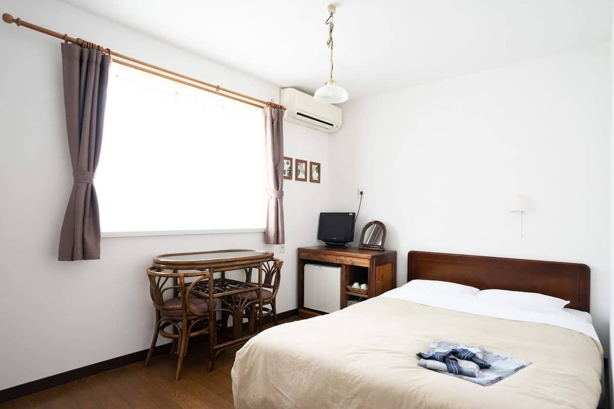Um quarto com cama, escrivaninha com cadeira, televisão e janela com cortinas.