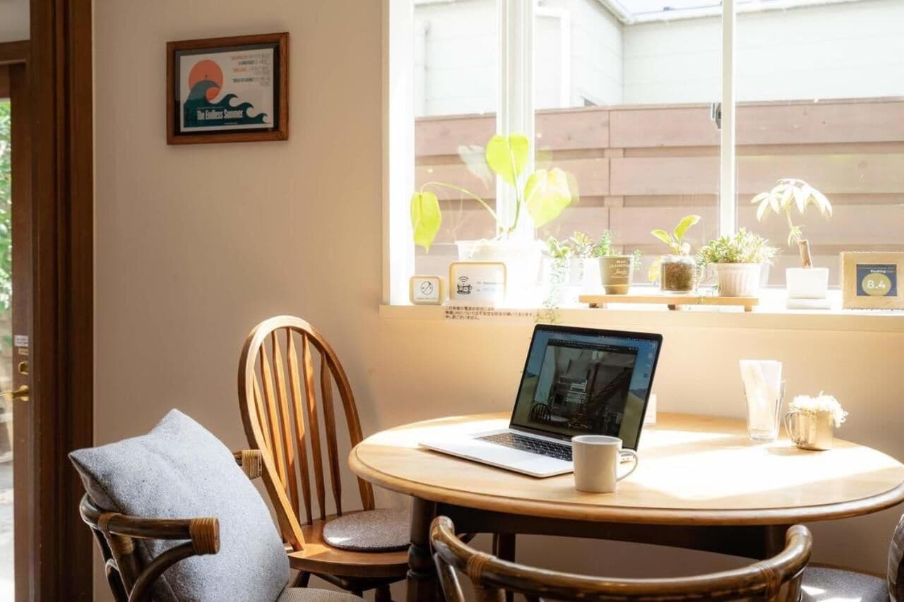 Uma cadeira de madeira com almofada, um laptop sobre uma mesa redonda pequena e algumas plantas em vasos no parapeito da janela.