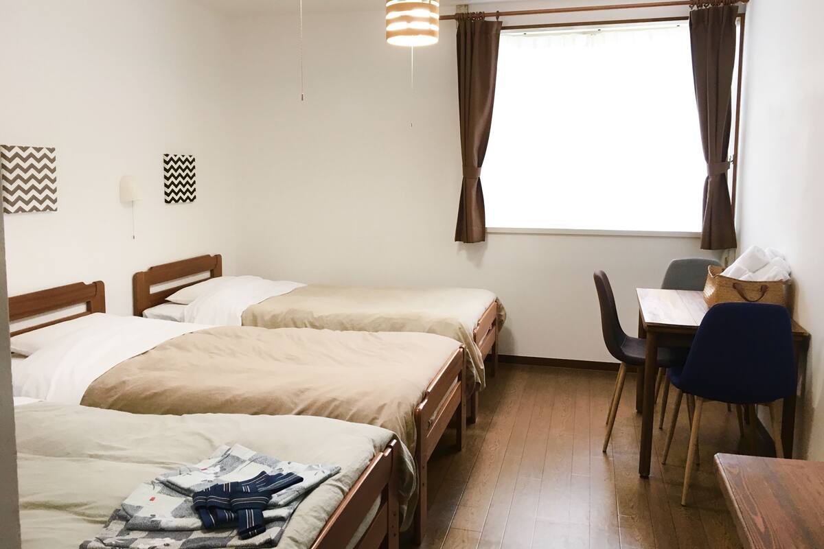 Quarto de hotel com duas camas de solteiro, uma escrivaninha com cadeira, janela com cortinas e luminárias fixadas na parede.