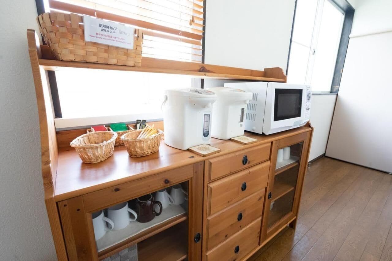 Uma cozinha compacta com micro-ondas, torradeira e cafeteira em uma bancada de madeira. Há cestos e uma placa indicando o uso de uma xícara usada.