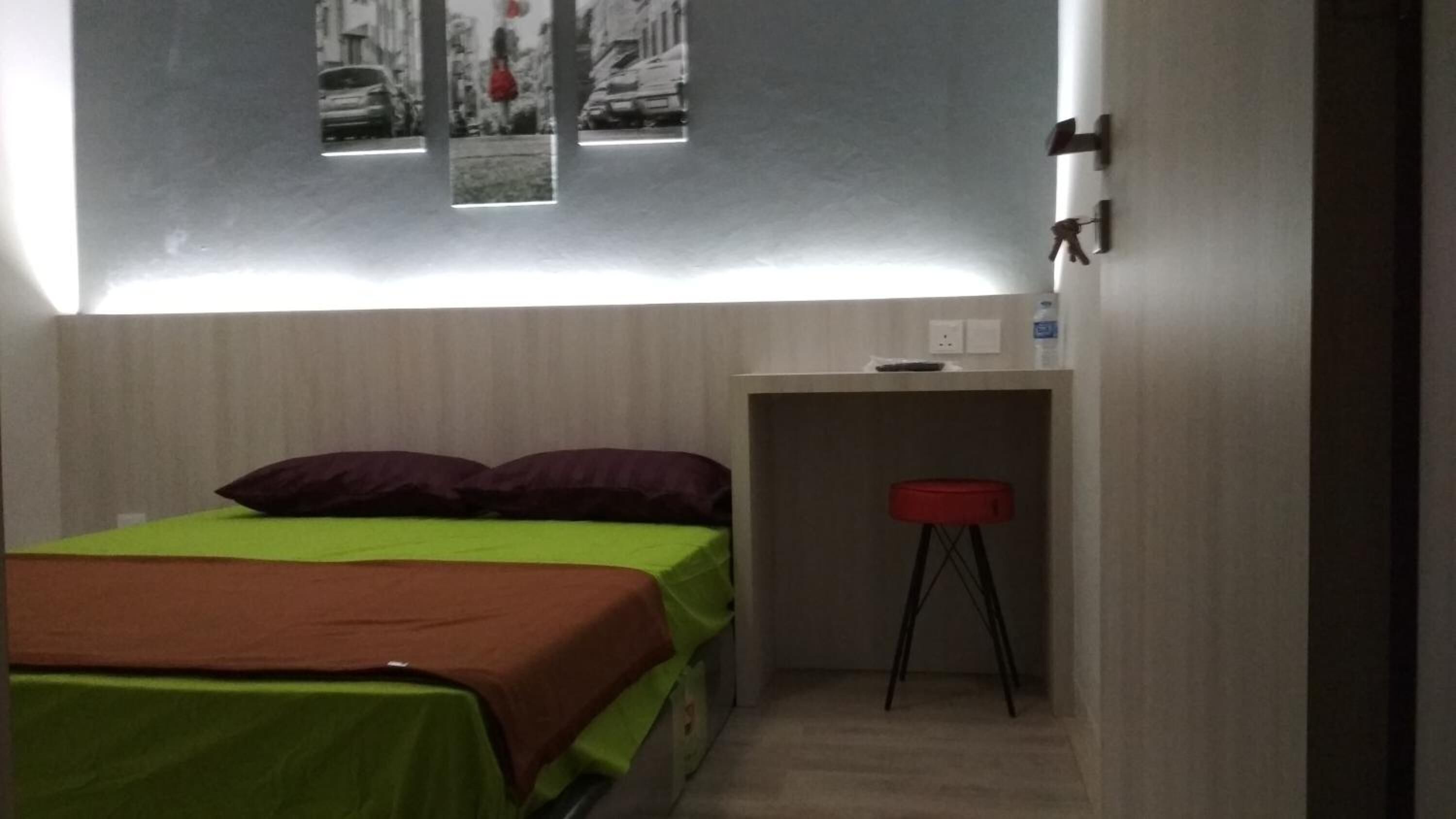 Um quarto pequeno e estreito com uma cama, um banquinho vermelho e uma prateleira fixada na parede.