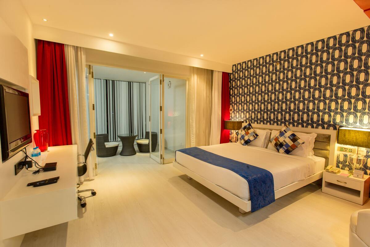 Quarto de hotel moderno com uma cama grande, uma escrivaninha com computador, uma televisão e um banheiro visível através de uma porta de vidro.