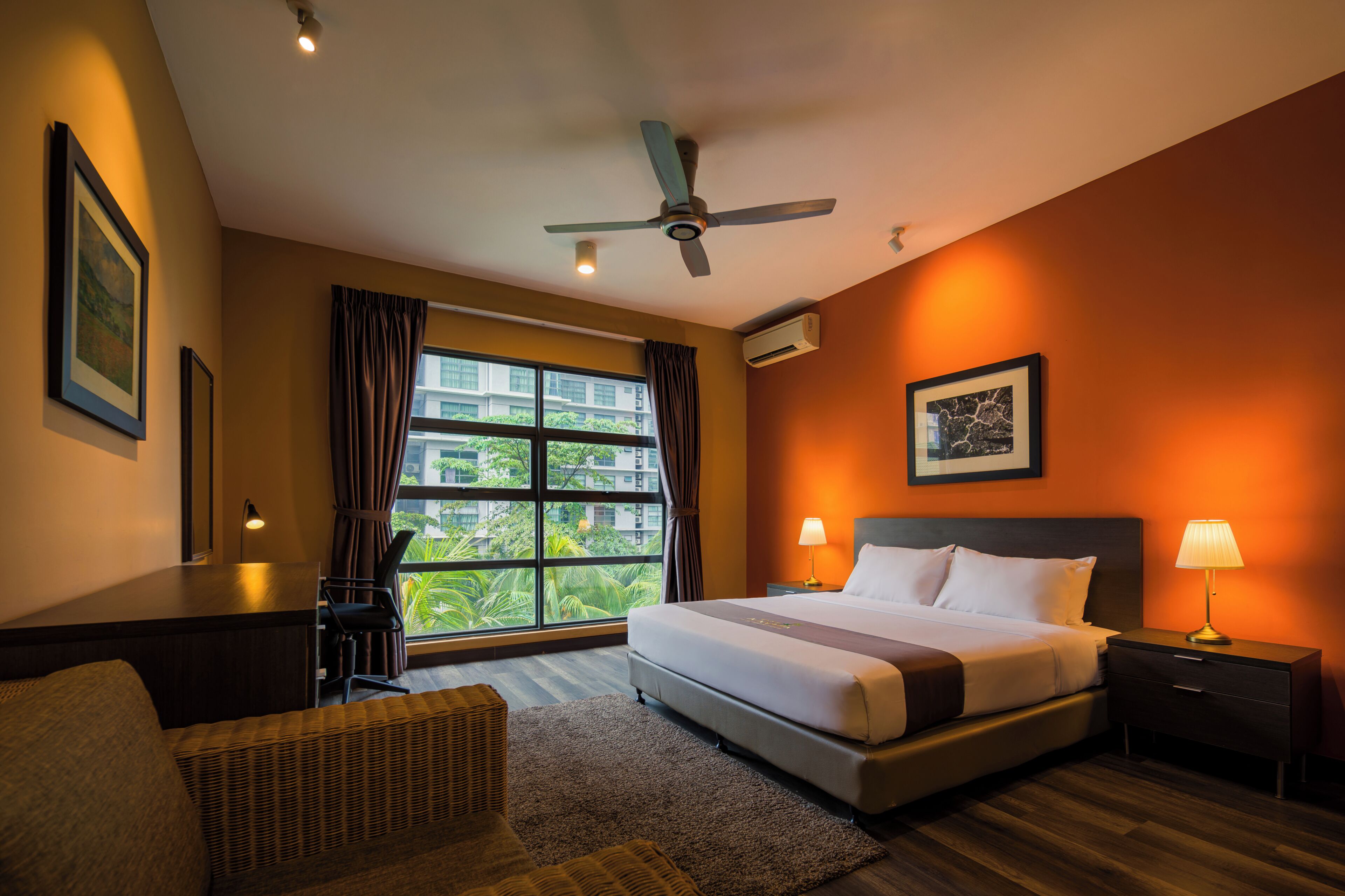 Quarto de hotel com cama, mesas de cabeceira, escrivaninha, cadeira, janela com cortinas, ventoinha no teto e quadros nas paredes.