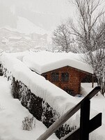 Uma cabana de madeira parcialmente coberta por neve, com uma janela visível e uma escadaria que sobe.