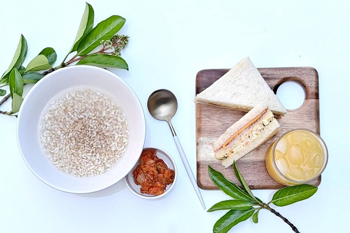 Uma tigela de arroz branco, um sanduíche em uma tábua de madeira, uma colher, um pequeno recipiente com molho vermelho e um copo com líquido amarelo.