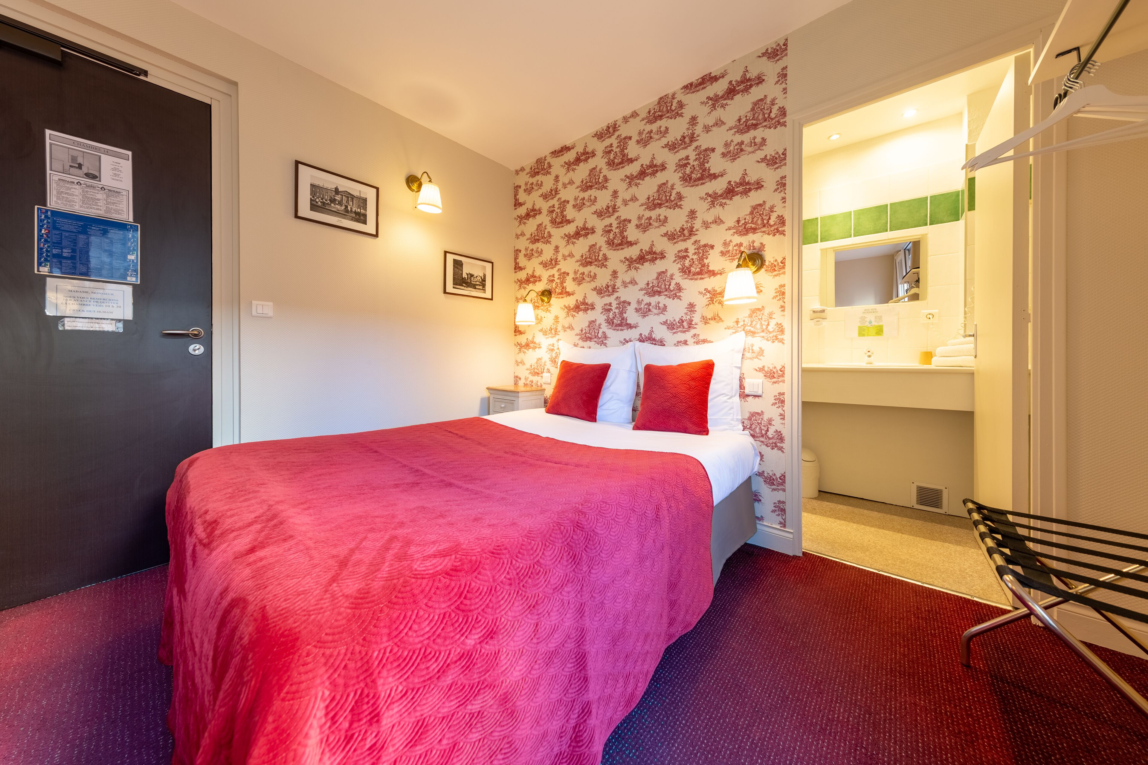 Quarto de hotel com cama, manta vermelha, tapete vermelho, parede com papel de parede floral e banheiro visível por uma porta aberta.