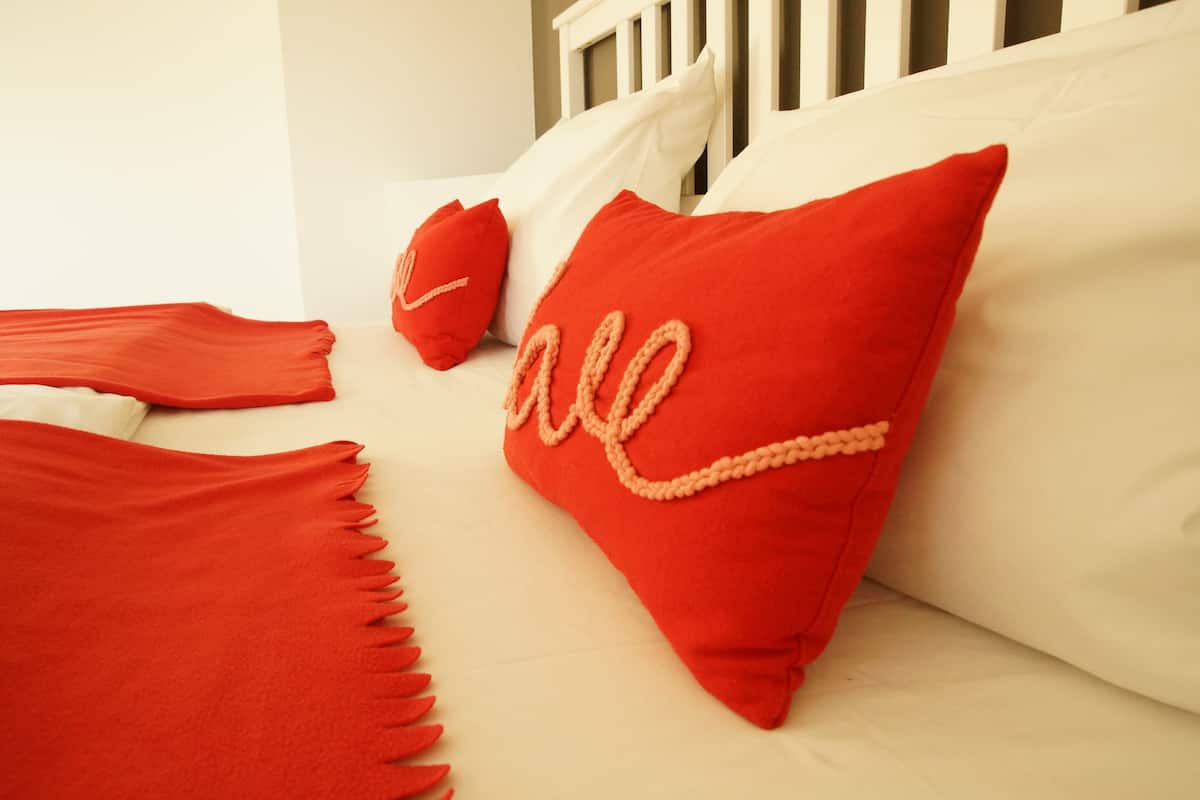 Cama com roupa de cama branca e almofadas vermelhas, uma delas com a palavra 'all' bordada.
