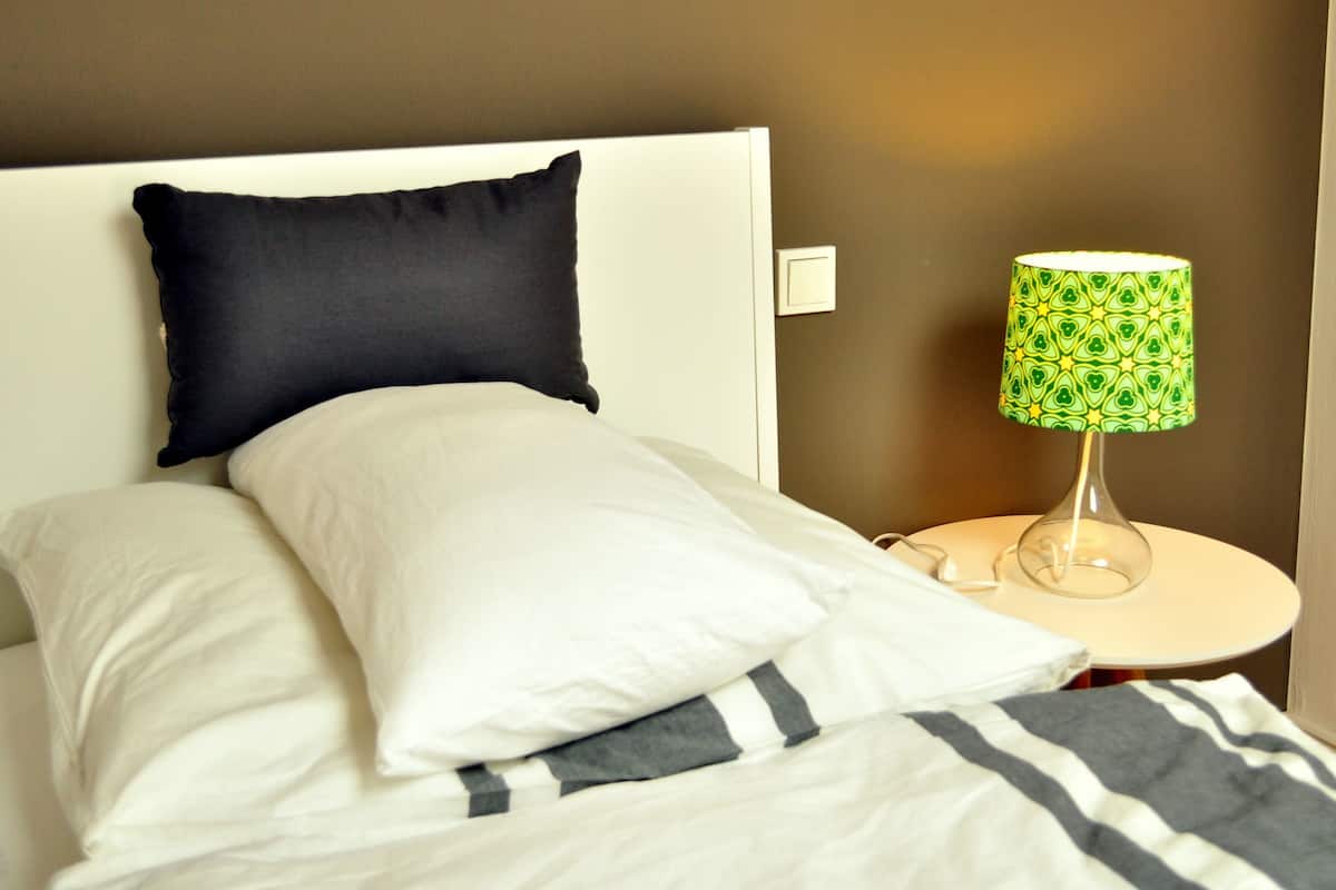 Uma cama bem arrumada com roupa de cama branca e preta, uma luminária estampada de verde sobre uma pequena mesa de cabeceira, e uma parede lisa ao fundo.