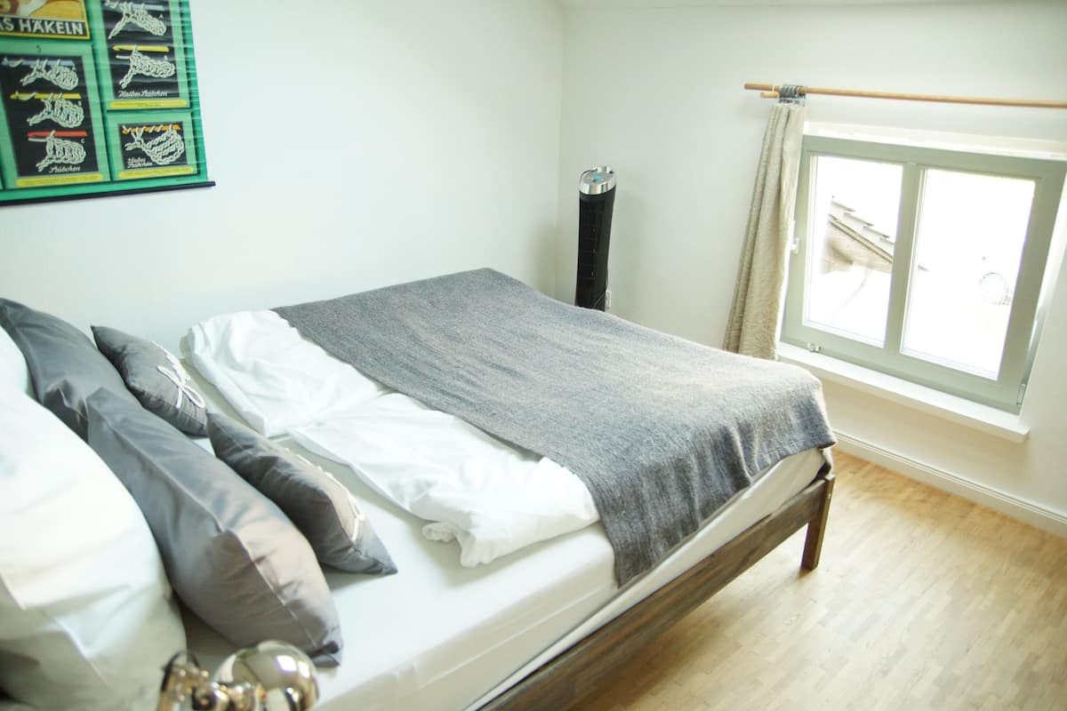 Uma cama bem arrumada com almofadas e um cobertor, piso de madeira, janela com cortinas e uma parede com quadros.