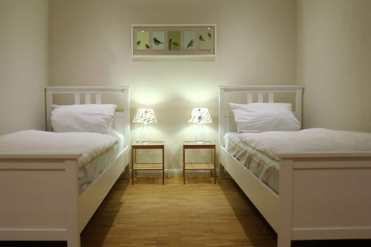 Quarto com duas camas de solteiro, piso de madeira e prateleira fixada na parede com quadros.